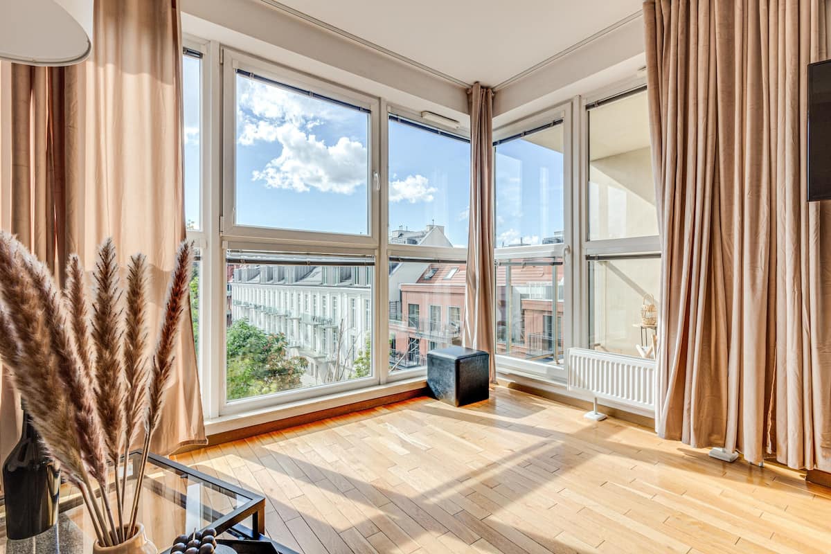 Quarto amplo com grandes janelas, um radiador e uma pequena mesa com objetos decorativos.