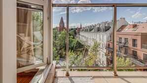 Quarto com amplas janelas que oferecem vista para a paisagem urbana, incluindo uma torre de igreja imponente e edifícios residenciais.