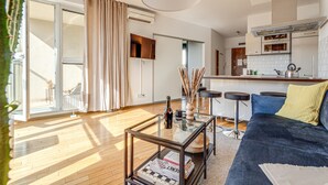 Uma sala de estar moderna com uma mesa de centro de vidro, um sofá azul-escuro e uma área de cozinha ao fundo.