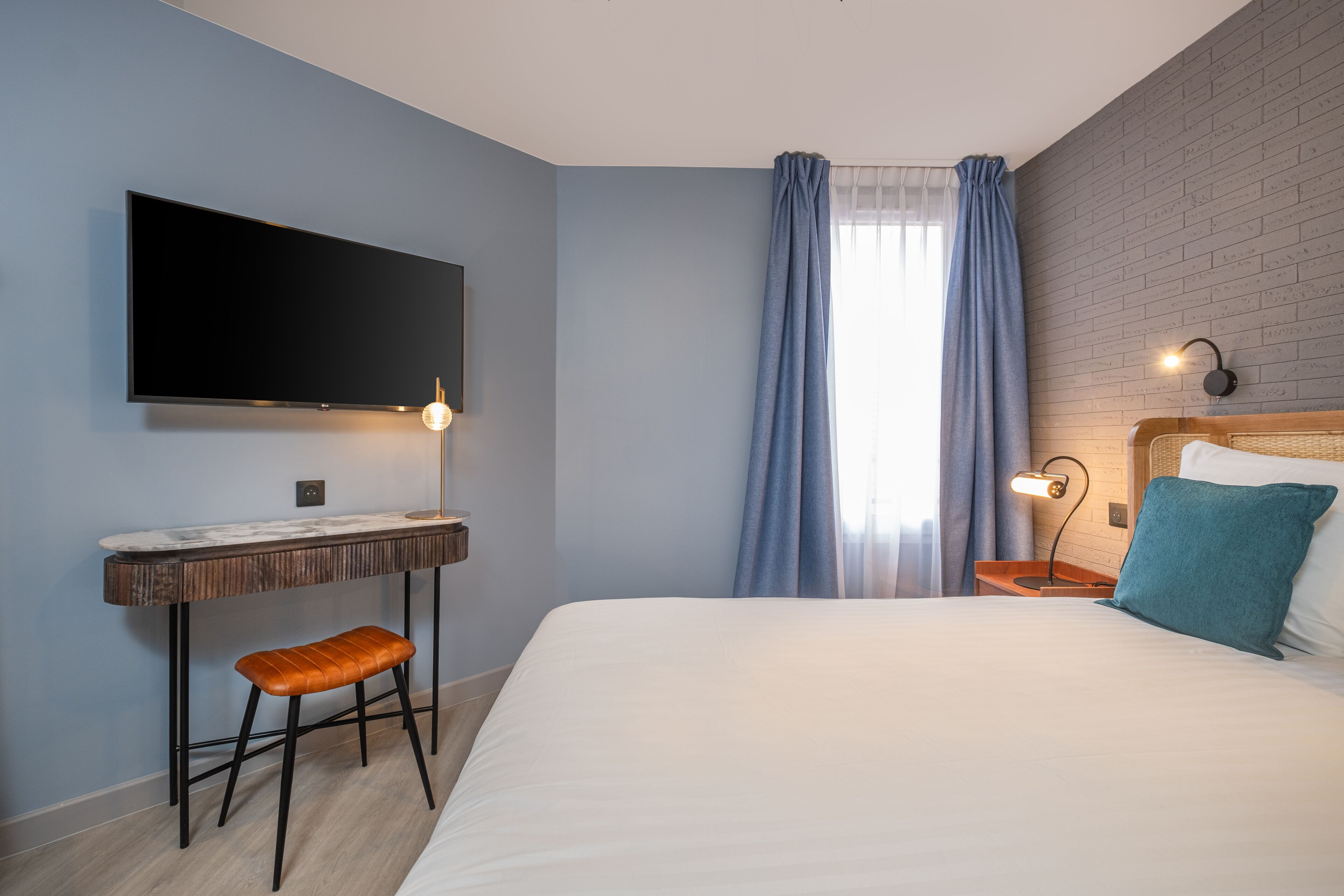 Quarto de hotel com cama, televisão fixada na parede, escrivaninha com cadeira e janela com cortinas.