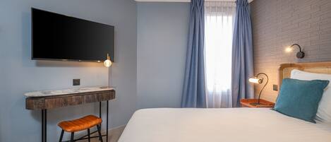 Quarto de hotel com cama, televisão fixada na parede, escrivaninha com cadeira e janela com cortinas.