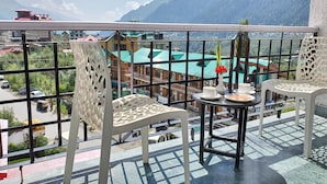 Varanda com duas cadeiras brancas, uma mesa pequena e um vaso com uma flor vermelha. A varanda tem vista para uma cidade com edifícios e uma montanha ao fundo.