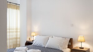 Cama bem arrumada com colcha cinza, duas luminárias de cabeceira e janela com cortinas.