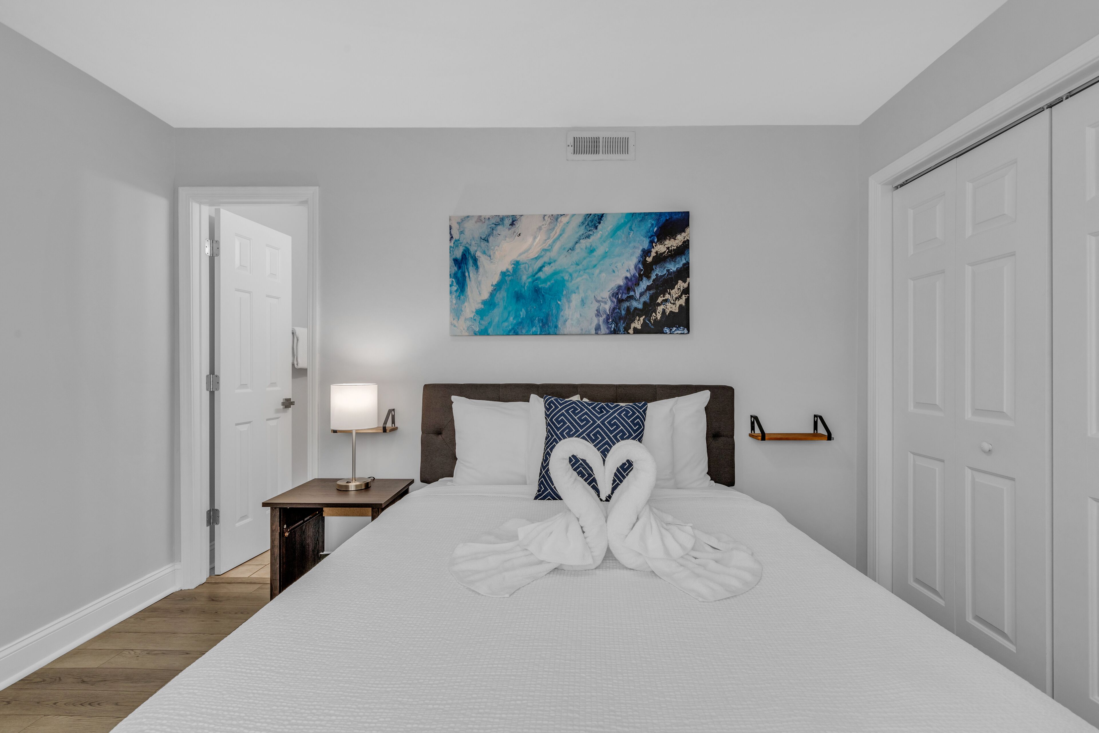 Quarto de hotel com cama, mesa de cabeceira, luminária, quadro e porta.