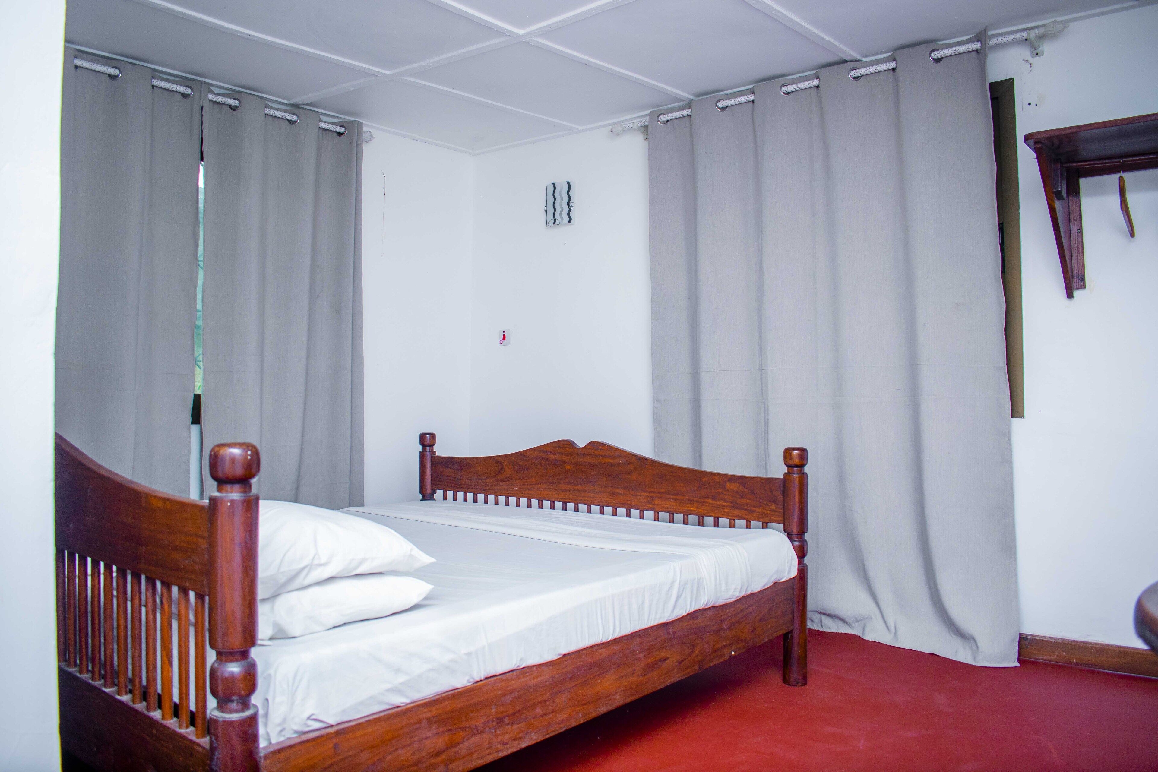 Cama de solteiro com estrutura de madeira e roupa de cama branca em quarto com paredes brancas e tapete vermelho.