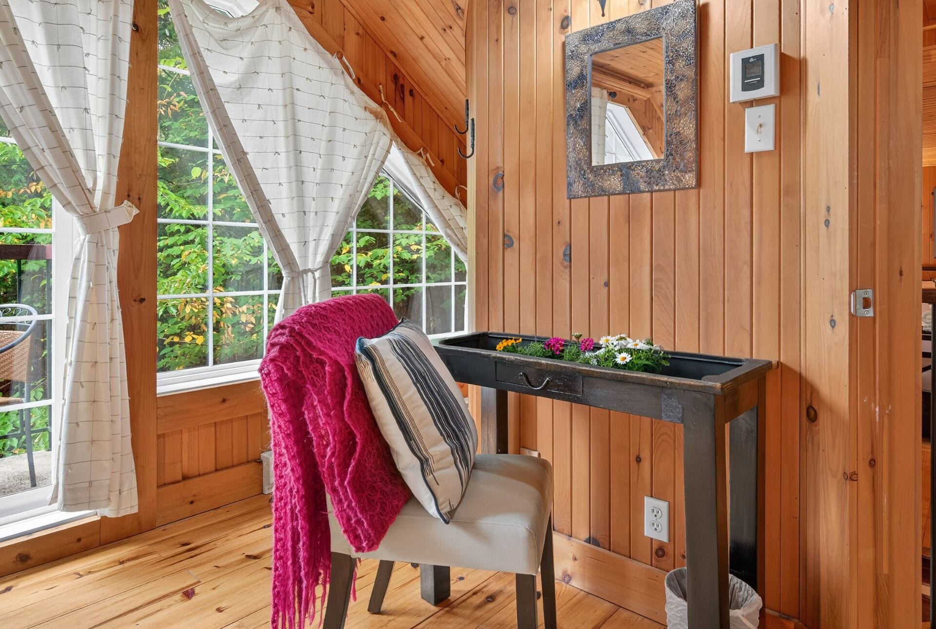 Quarto aconchegante com paredes de madeira, janela com cortinas, um banco com manta rosa, mesa de cabeceira com espelho e uma pequena mesa de cabeceira com flores.