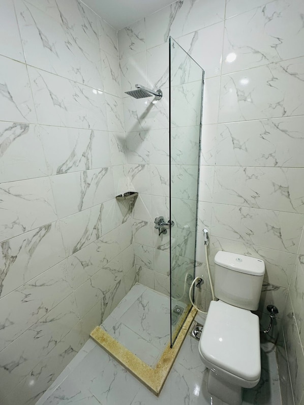 Casa de banho com chuveiro em cabine de vidro, azulejos de mármore branco e sanita branca.