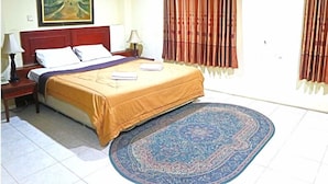 Quarto de hotel com cama, mesa de cabeceira, luminária, pintura na parede, janela com cortinas, aparelho de ar condicionado e tapete estampado.