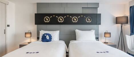 Quarto de hotel com duas camas, uma cabeceira moderna, luminárias laterais e um ponto de luz fixado na parede.