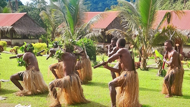 Um grupo de pessoas vestidas com trajes típicos sentadas em bancos de palha em uma área gramada, com palmeiras e montanhas ao fundo.