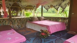 Quarto com duas camas, cada uma com colcha floral rosa e uma almofada. Uma pequena mesa com um vaso de flores está no centro. O quarto tem um telhado de palha e vista para um jardim exuberante lá fora.