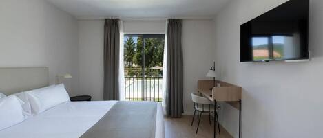 Quarto de hotel com uma cama grande, uma escrivaninha com cadeira, uma televisão na parede e vista para o exterior através de uma janela.