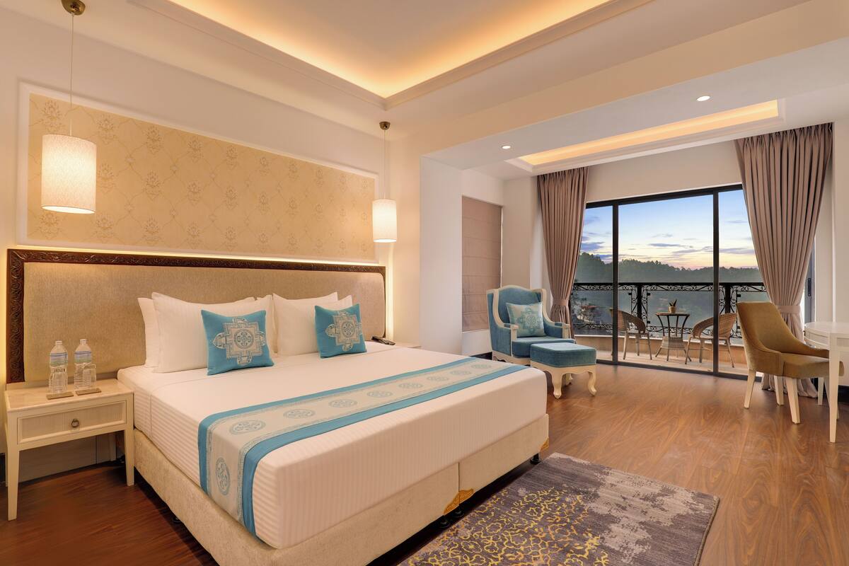 Quarto de hotel com uma cama grande, mesa de cabeceira, uma cadeira, uma pequena mesa e vista para o exterior.