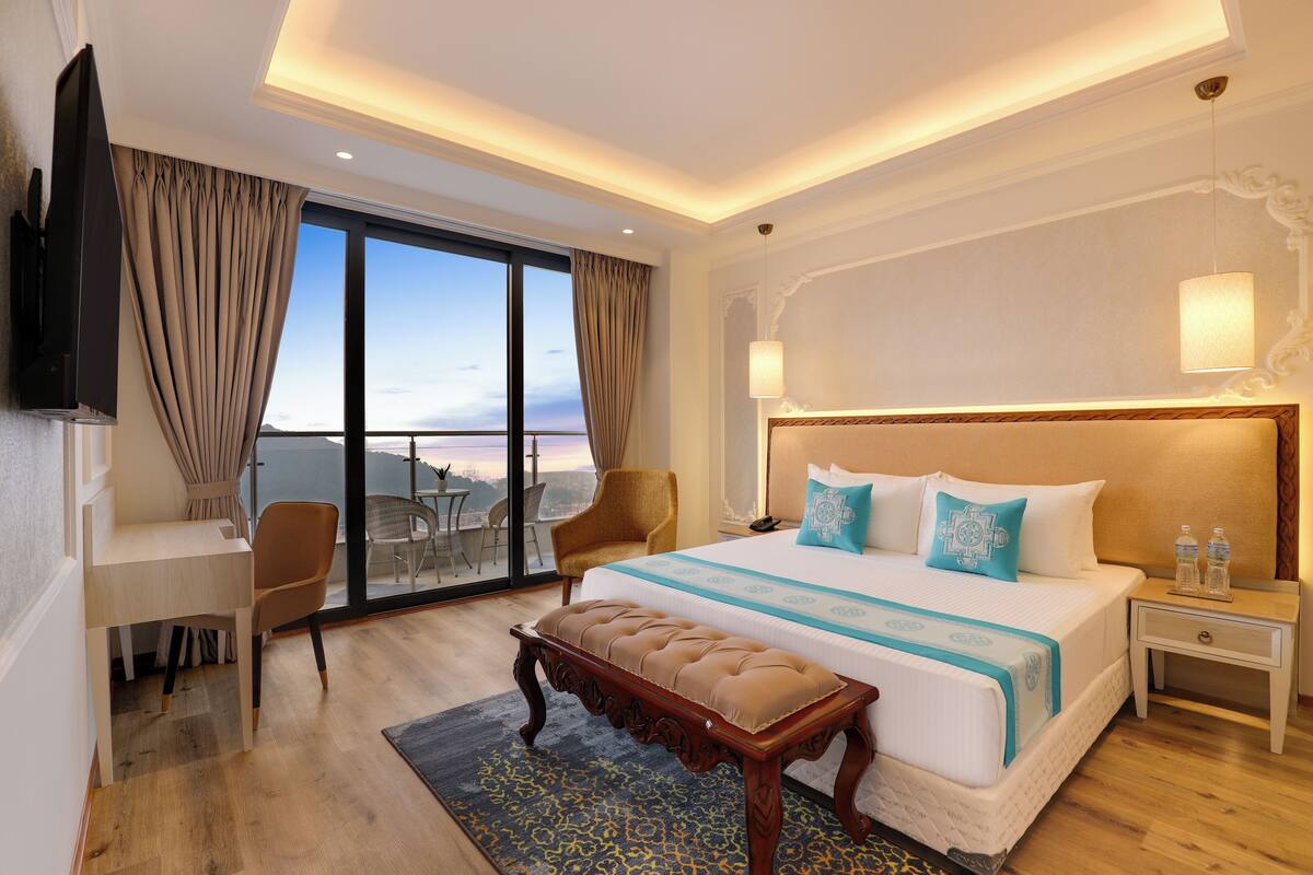 Quarto de hotel com uma cama grande, uma escrivaninha, uma cadeira, um banco, uma televisão e vista para a cidade.