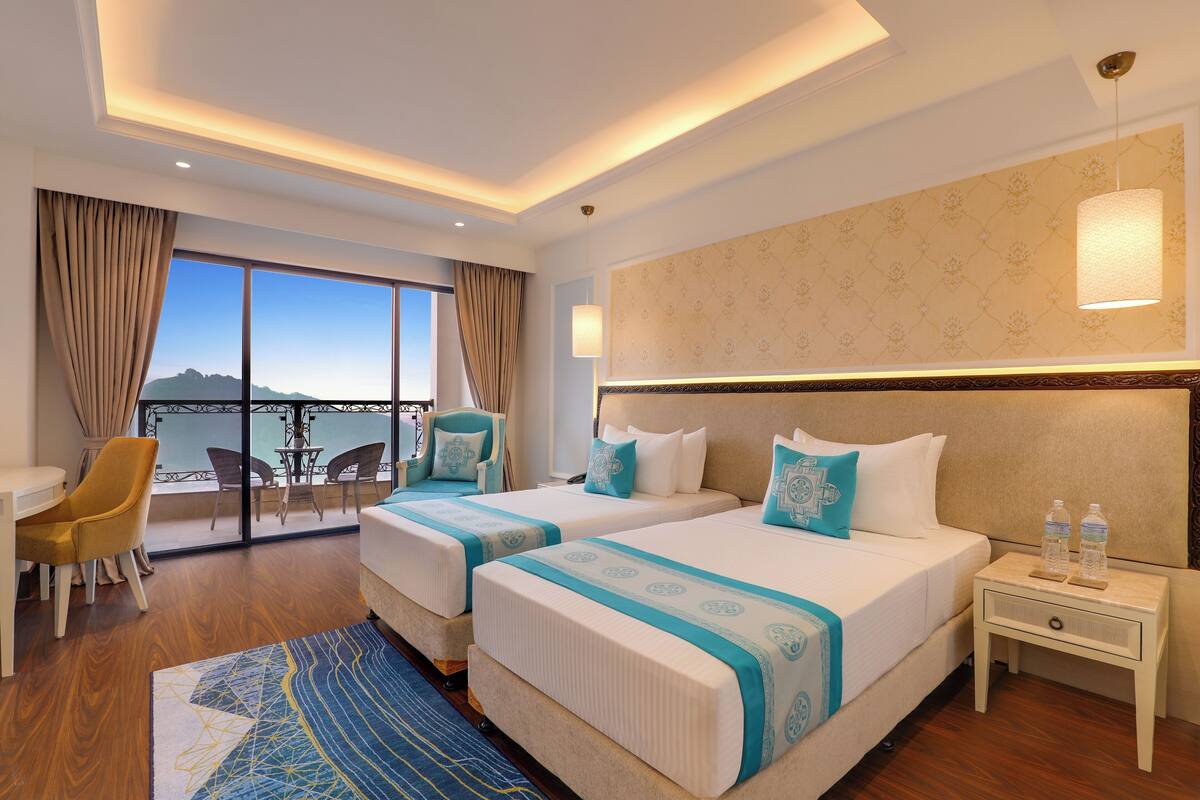 Quarto de hotel com duas camas, varanda com vista, área de refeições e mesa de cabeceira com garrafas de água.
