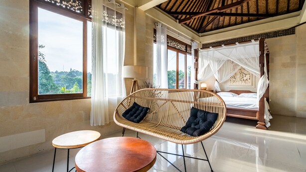 Quarto com cama, cadeira de vime, mesa redonda e janelas amplas.