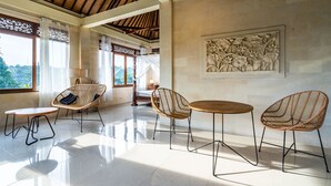 Um quarto amplo com teto alto, piso de azulejos polidos e uma grande janela com vista para o verde. Há cadeiras de vime e uma mesa redonda de madeira.