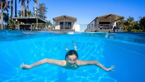 Piscina com água azul cristalina, rodeada por palmeiras e dois edifícios com áreas cobertas.