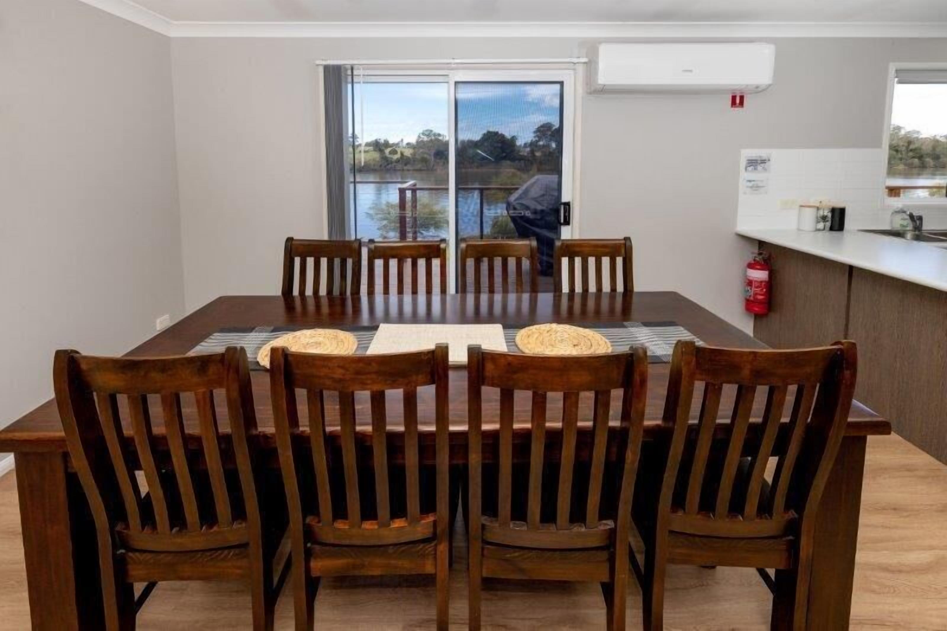 Uma área de jantar com uma mesa grande e seis cadeiras de madeira, uma porta de vidro deslizante que dá para uma vista de um corpo d'água, e uma cozinha com bancada branca e um extintor de incêndio.