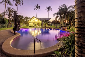 Piscina com borda curva, rodeada por palmeiras e um edifício com telhado iluminado.