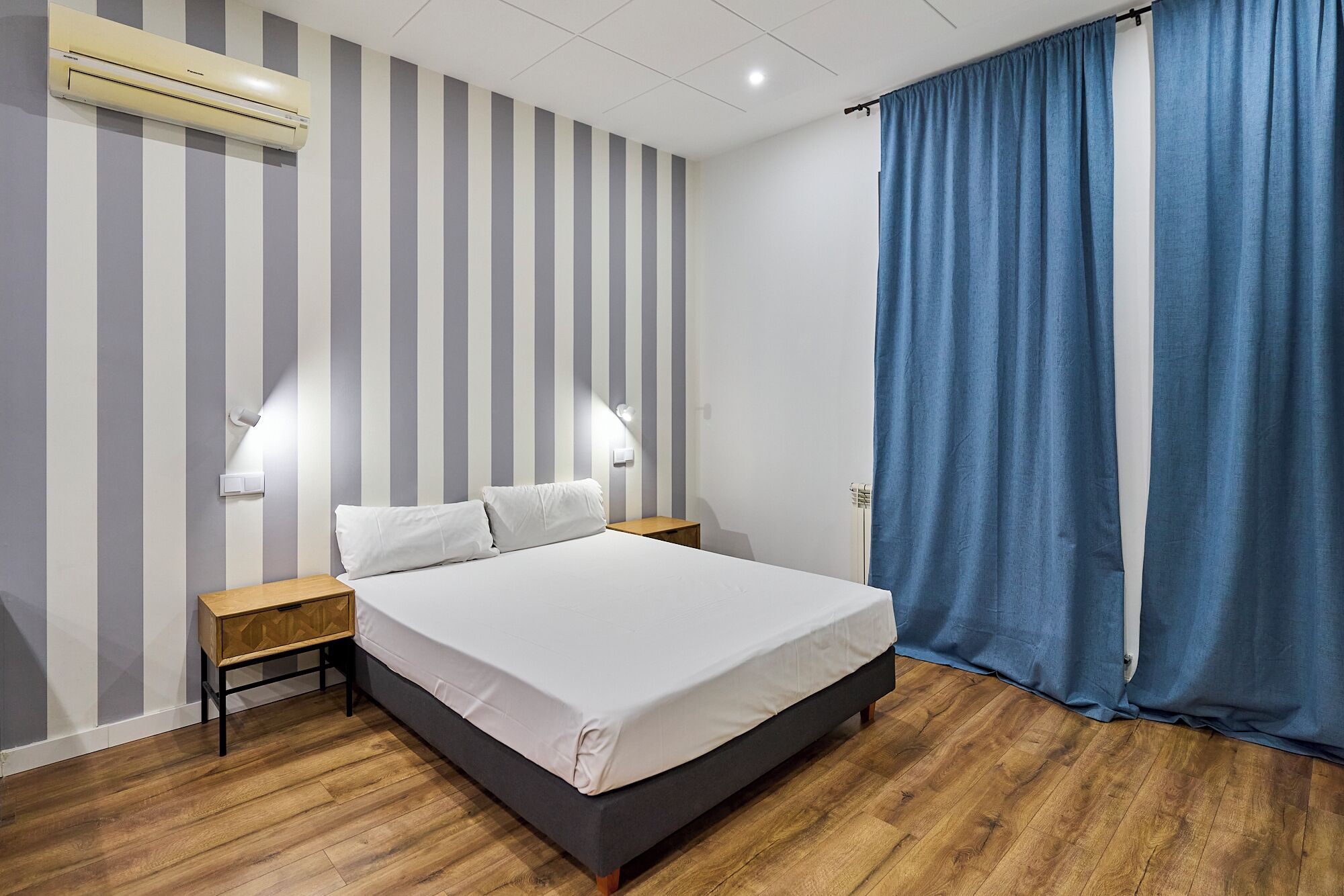 Quarto com cama, piso de madeira, parede listrada, ar condicionado, abajures e cortina azul.
