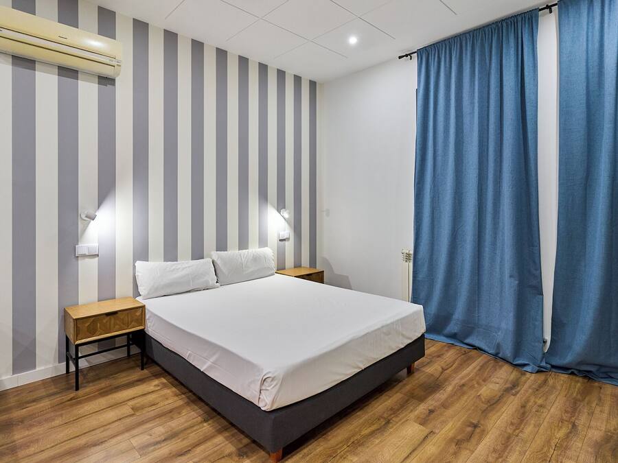 Quarto com cama, piso de madeira, parede listrada, ar condicionado, abajures e cortina azul.