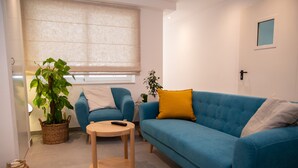 Uma sala de estar moderna com um sofá azul, uma mesinha de madeira e duas cadeiras. Há plantas em vasos e uma janela com persianas.