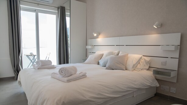 Quarto de hotel moderno com uma cama grande, uma mesinha de cabeceira e uma vista através de uma janela ampla.