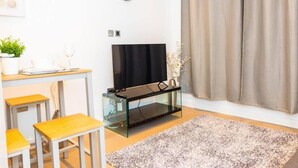 Uma sala de estar moderna com televisão de tela plana em suporte de vidro, uma mesa de jantar com cadeiras, uma pequena planta em vaso e uma janela grande com cortinas.