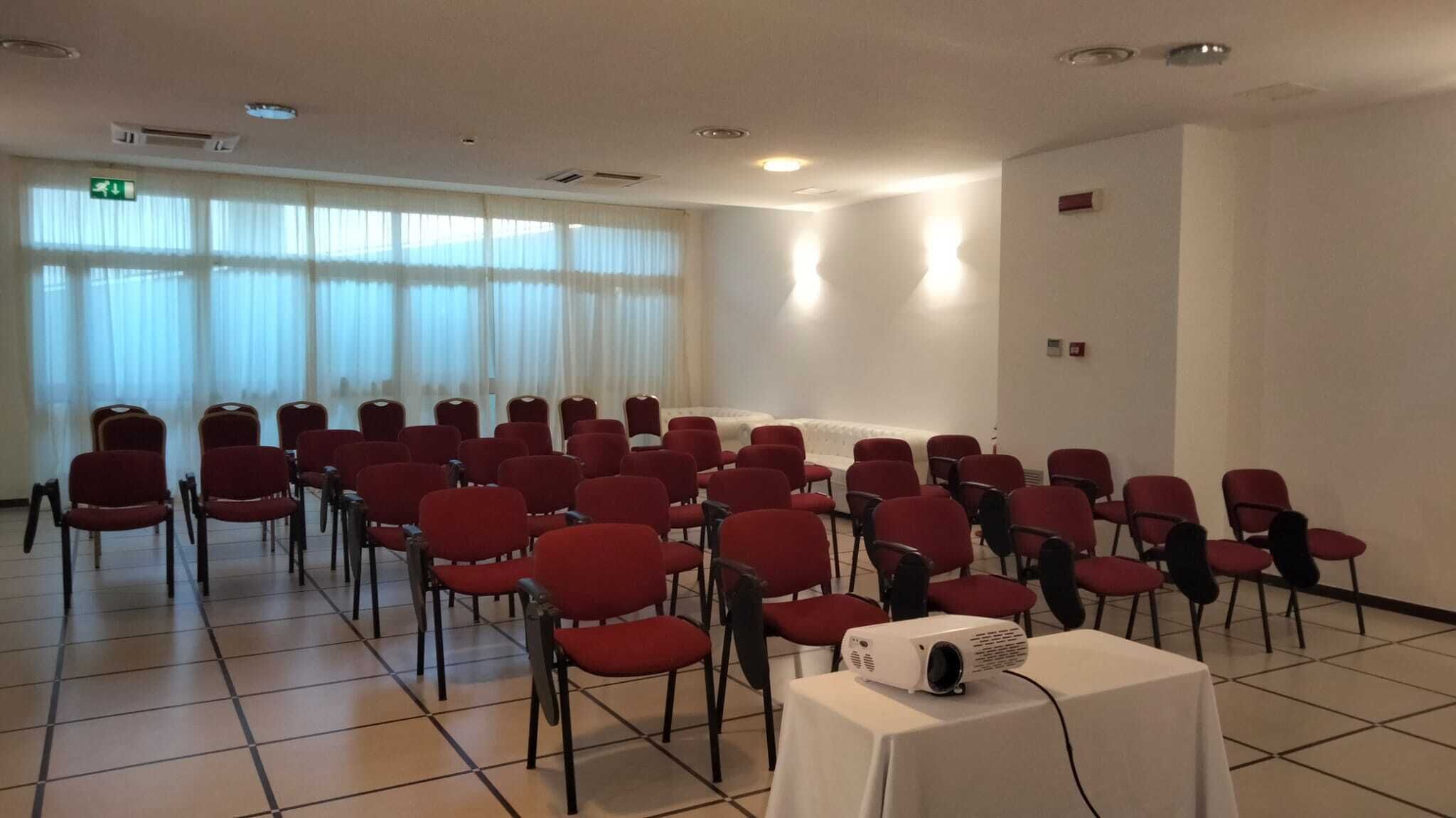 Uma sala de conferências com fileiras de cadeiras vermelhas, um projetor sobre uma mesa e grandes janelas com cortinas.