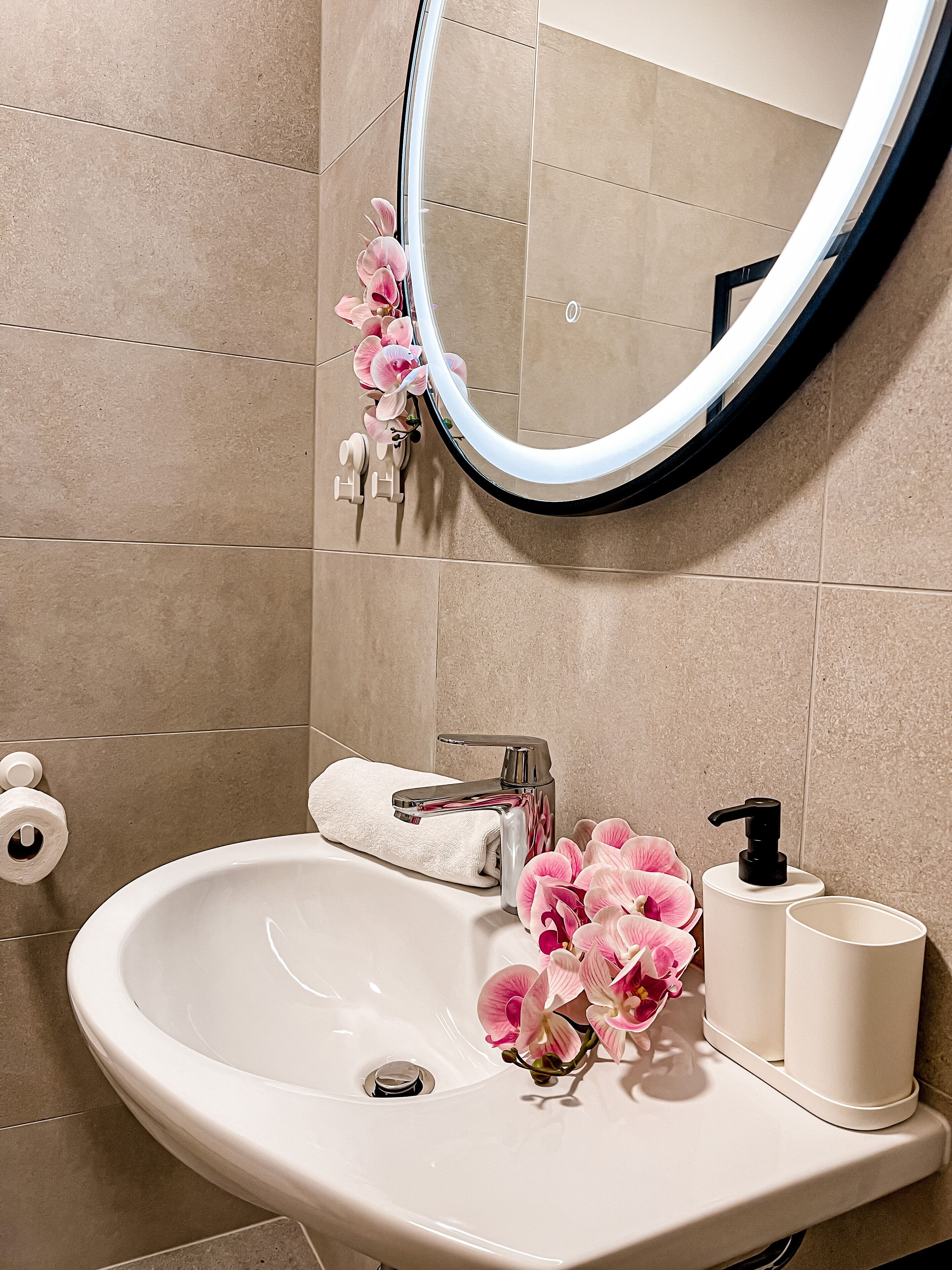 Casa de banho com uma pia branca, um espelho, uma toalha, porta-papel higiénico e flores cor-de-rosa.