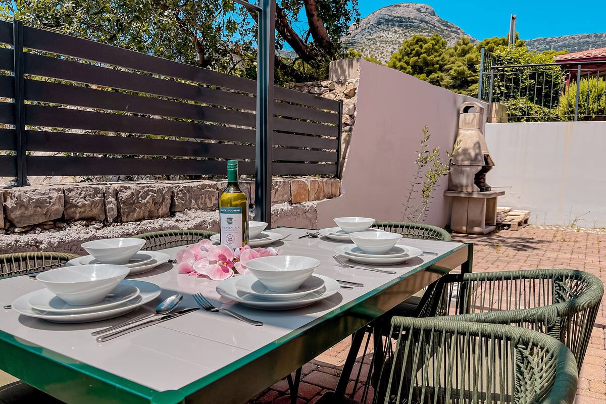 Mesa posta para quatro com pratos, tigelas e talheres brancos, uma garrafa de vinho e flores cor-de-rosa sobre uma mesa de vidro em ambiente externo, com uma parede de pedra e vegetação ao fundo.