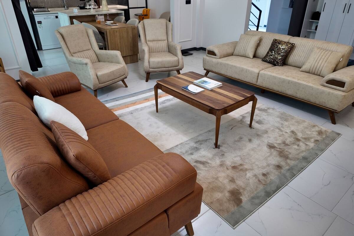 Uma sala de estar com um sofá marrom, poltronas bege, um sofá bege com almofada dourada e uma mesa de centro de madeira.