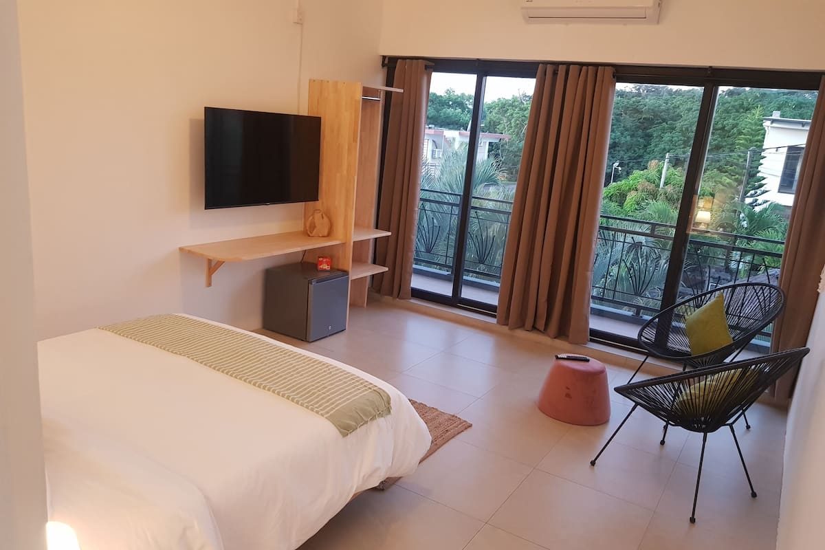 Quarto de hotel moderno com cama, TV, mini-geladeira, cadeira e varanda com vista para área verde.