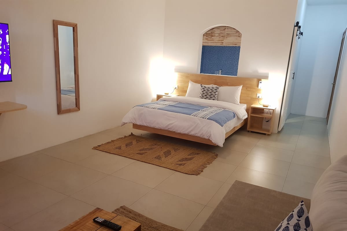 Quarto com cama, mesas de cabeceira, um criado-mudo, televisão, espelho e porta.