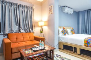 Quarto de hotel com sofá, uma mesinha com bandeja de lanches, cama com almofadas coloridas e um ar condicionado.
