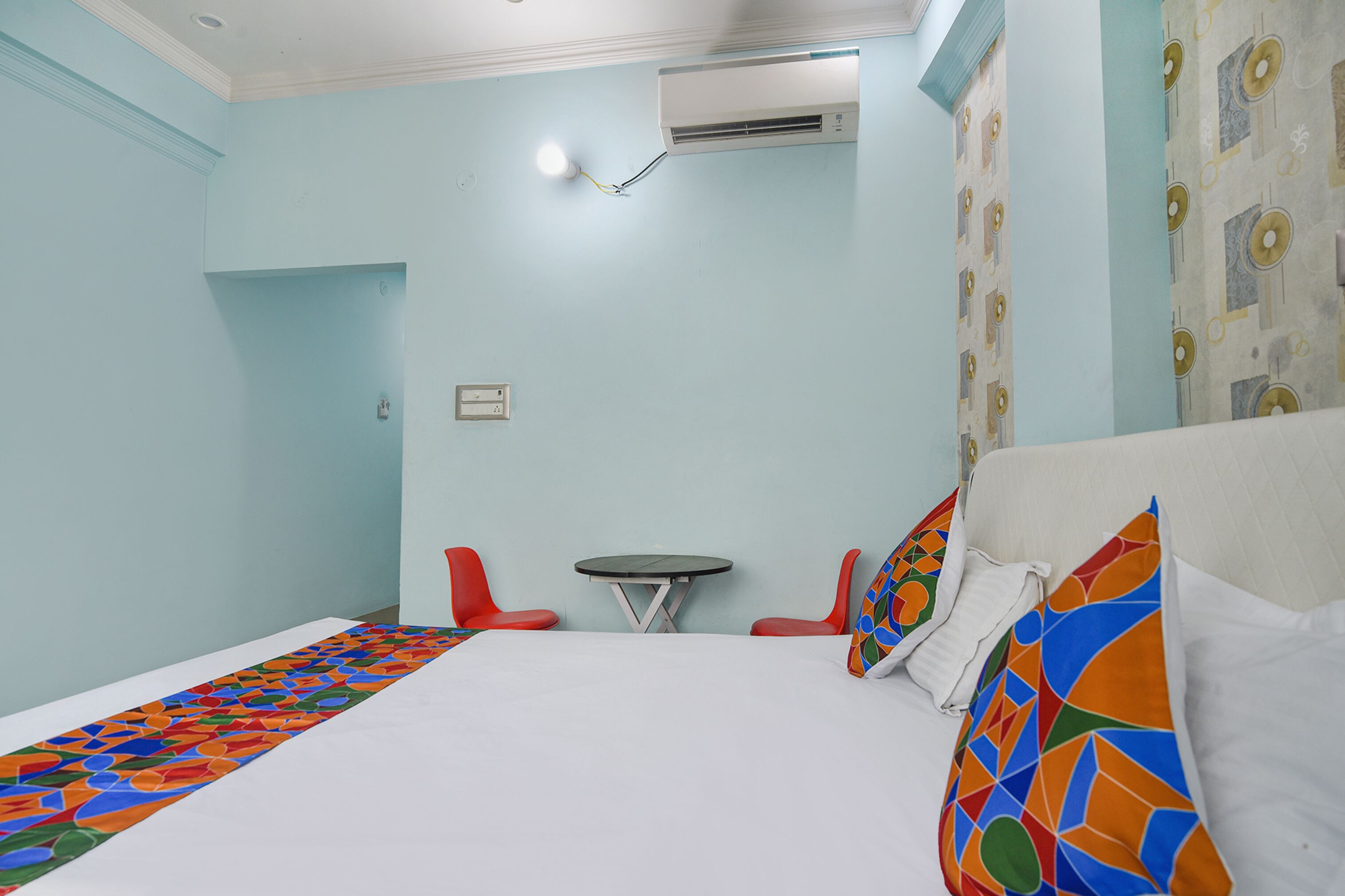 Quarto de hotel com uma cama, uma mesinha, duas cadeiras vermelhas e um ar condicionado.