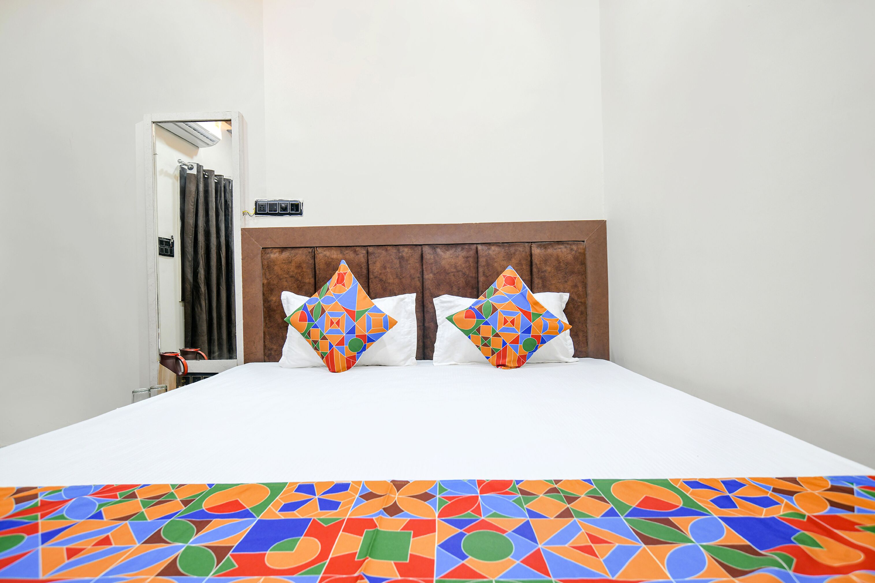 Quarto de hotel com uma cama grande, duas almofadas coloridas em padrão geométrico, uma cabeceira de madeira e uma parede branca lisa.