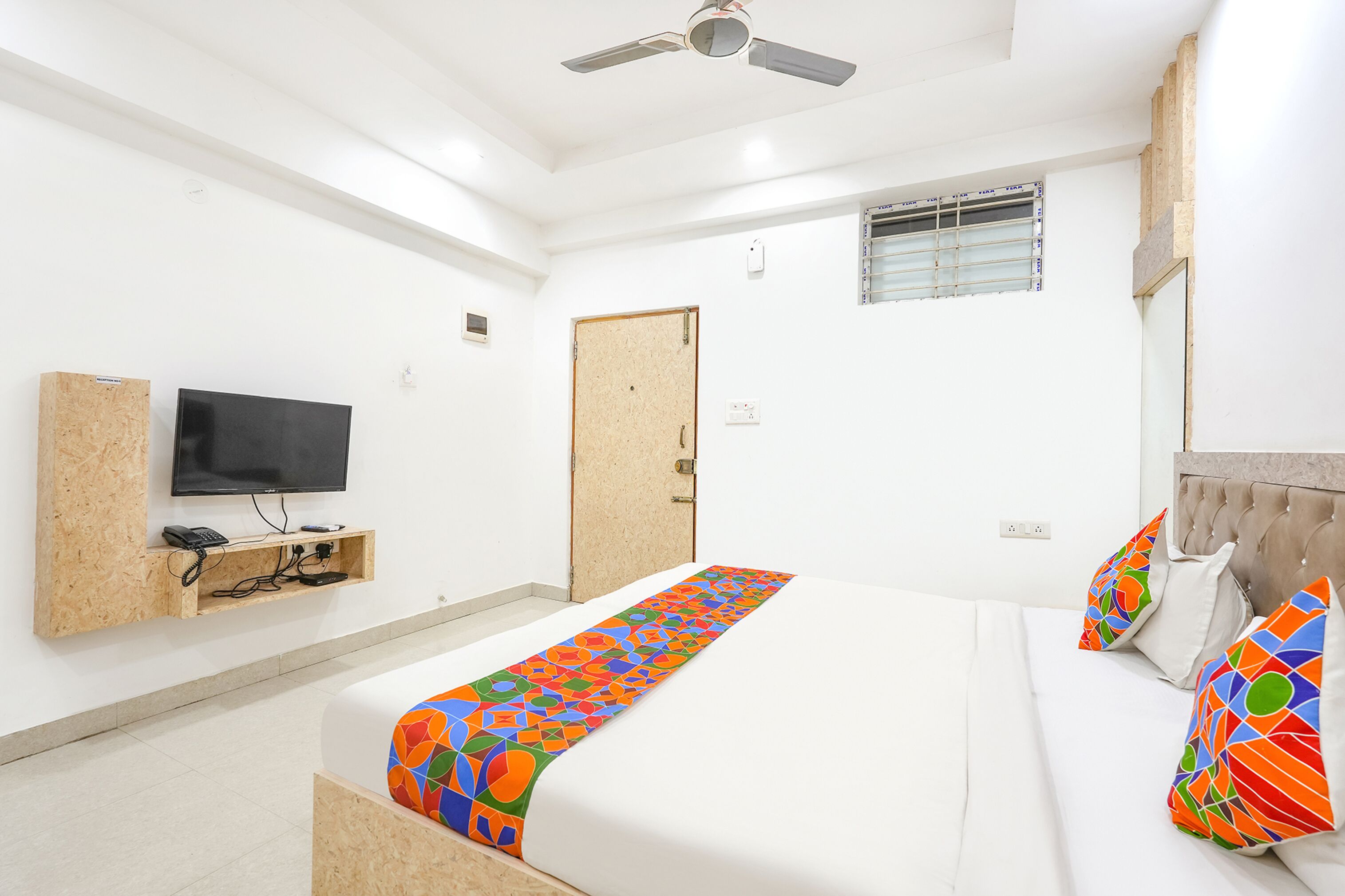 Quarto de hotel com cama, televisão, prateleira de madeira e ventoinha no teto.