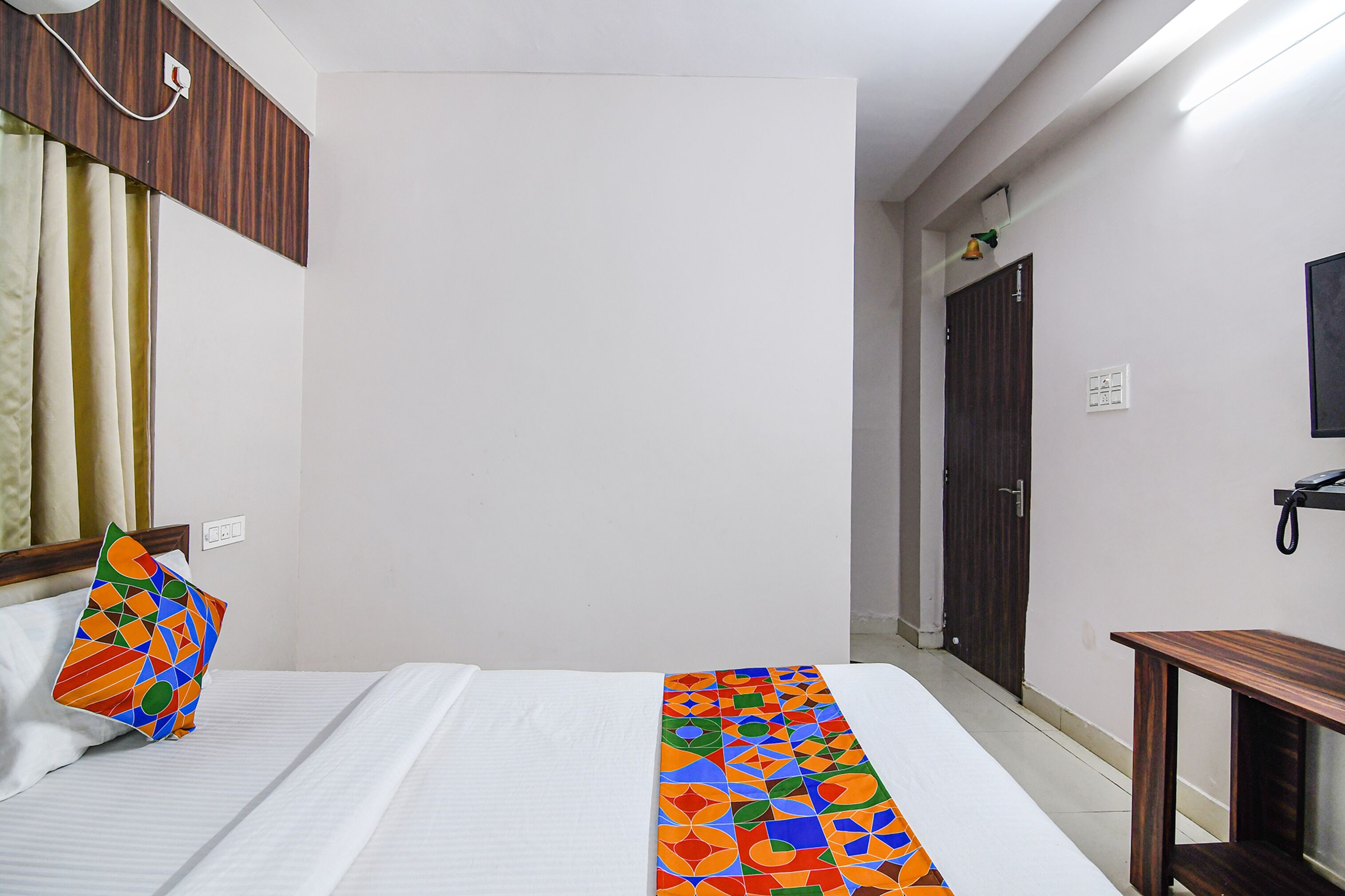 Quarto de hotel com cama, cobre de cama colorido, cabeceira de madeira, mesa de cabeceira e uma porta.