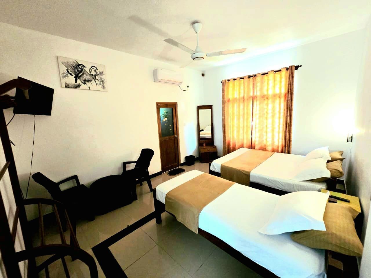 Quarto de hotel com duas camas, uma escrivaninha, uma cadeira, um ventilador de teto e uma janela com cortinas.