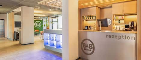Uma área de recepção de hotel com design moderno, apresentando um balcão de atendimento, uma parede verde com o logotipo do hotel e um espaço de estar com uma planta.
