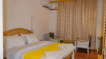 Quarto de hotel com cama, duas cadeiras, uma mesinha, aparelho de ar condicionado e cortina.