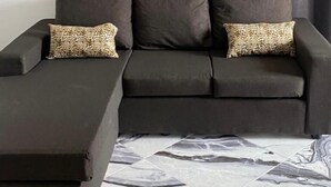 Um sofá de cor escura com três almofadas e um banquinho combinando, posicionado em frente a uma parede com cortina.
