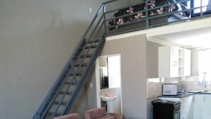 Uma sala de estar com sofá seccional, tapete estampado, escada e área de cozinha ao fundo.