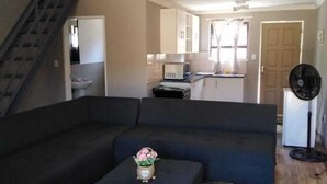 Sala de estar com um sofá escuro, um pequeno banquinho e um vaso decorativo. Uma escada leva a um quarto no mezanino acima.