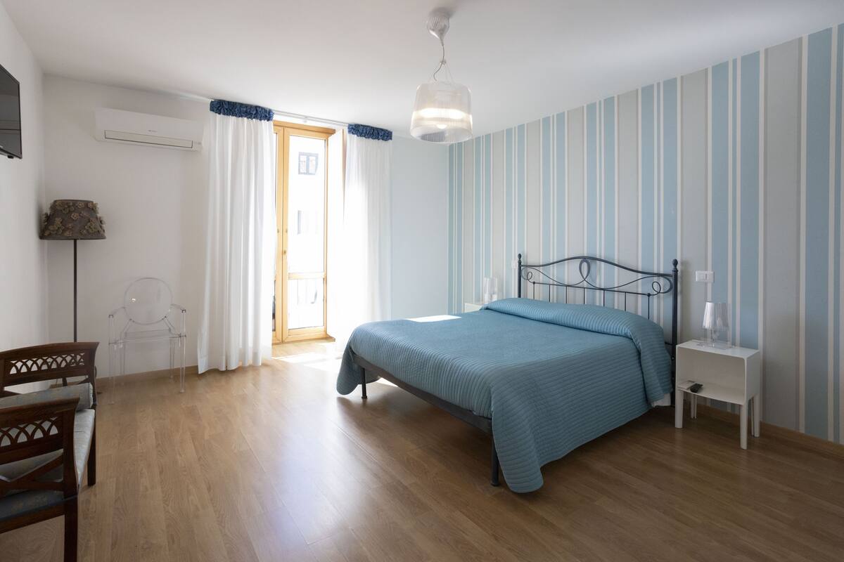 Quarto com cama, mesa de cabeceira, luminária, cadeira e janela com cortinas.