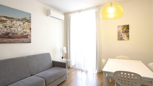 Uma sala de estar moderna com um sofá cinzento, uma mesa de jantar branca com cadeiras, uma luminária pendente e duas pinturas nas paredes.