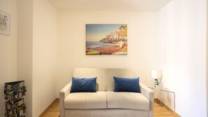 Quarto com sofá bege, duas almofadas azuis, pintura de uma paisagem costeira, mesa pequena com abajur e estante de livros.