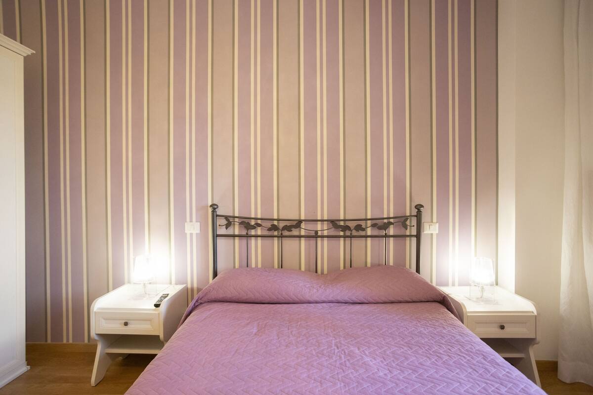 Uma cama bem arrumada com colcha rosa, duas mesas de cabeceira com abajures e uma parede listrada ao fundo.